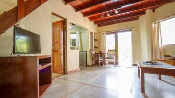 Uma sala de estar ampla com teto de madeira, uma televisão sobre um suporte de madeira, uma mesa de jantar com cadeiras e uma cozinha visível através de uma porta aberta.