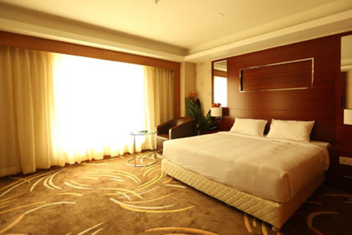Quarto de hotel com uma cama grande, uma escrivaninha, uma cadeira e um amplo janelão com cortinas.