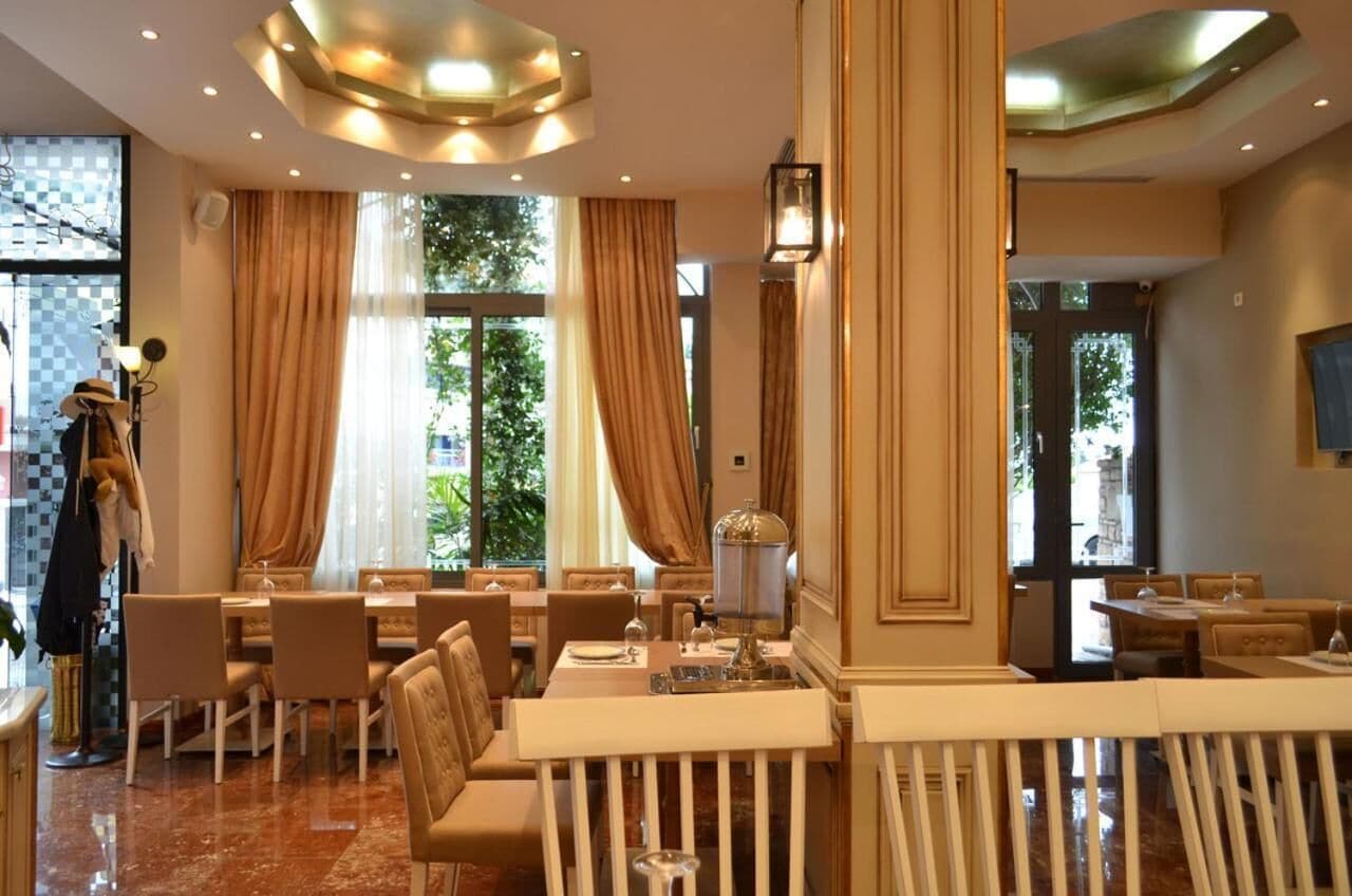 Um restaurante com teto alto, lustres e grandes janelas com cortinas. Mesas e cadeiras dispostas para refeições.
