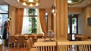 Um restaurante com teto alto, lustres e grandes janelas com cortinas. Mesas e cadeiras dispostas para refeições.