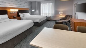 Quarto de hotel com duas camas, uma mesa, uma cadeira, uma luminária e uma janela grande com cortinas.