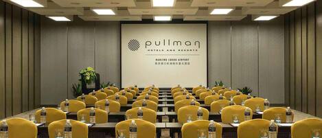 Uma sala de conferência com cadeiras, mesas e um grande ecrã exibindo a marca 'Pullman Hotels and Resorts'.