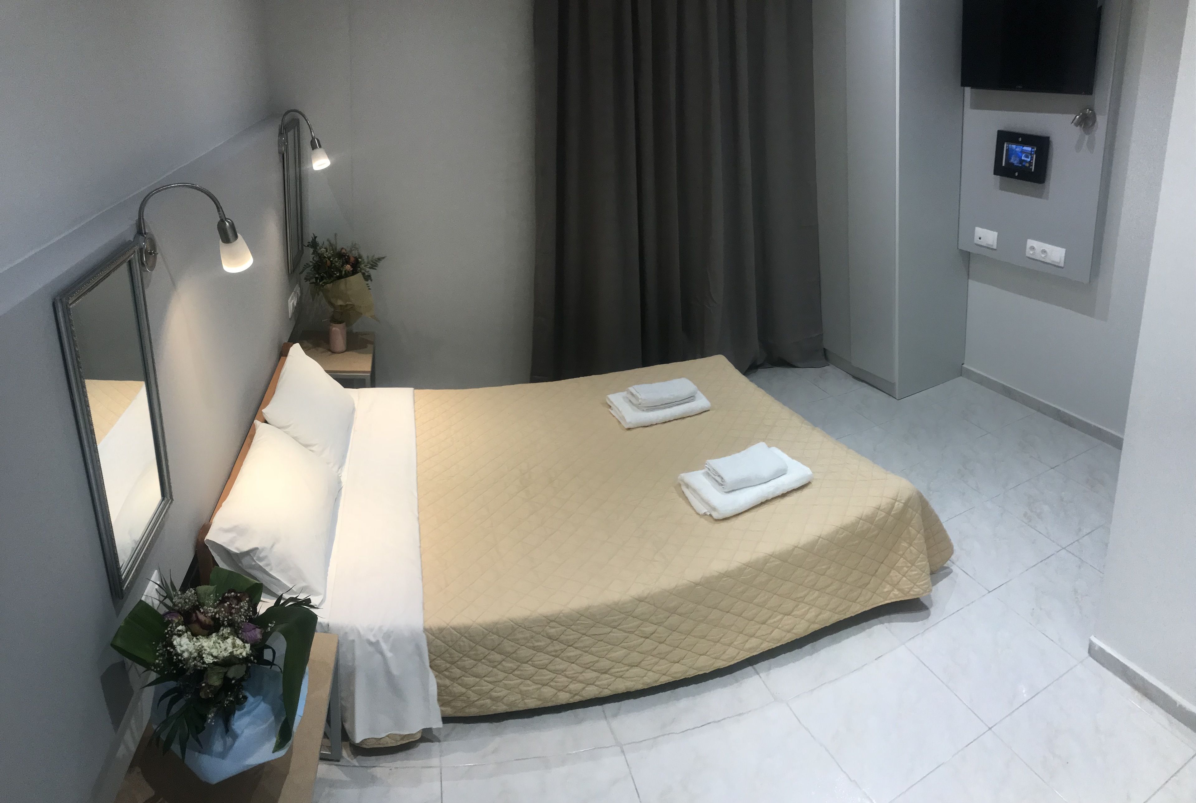 Cama bem arrumada com colcha bege, duas toalhas brancas, mesa de cabeceira com abajur, espelho e TV fixada na parede.