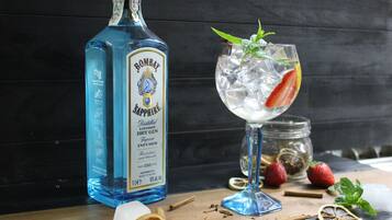 Uma garrafa de gin Bombay Sapphire e um copo com morango e folha de hortelã sobre uma mesa de madeira.