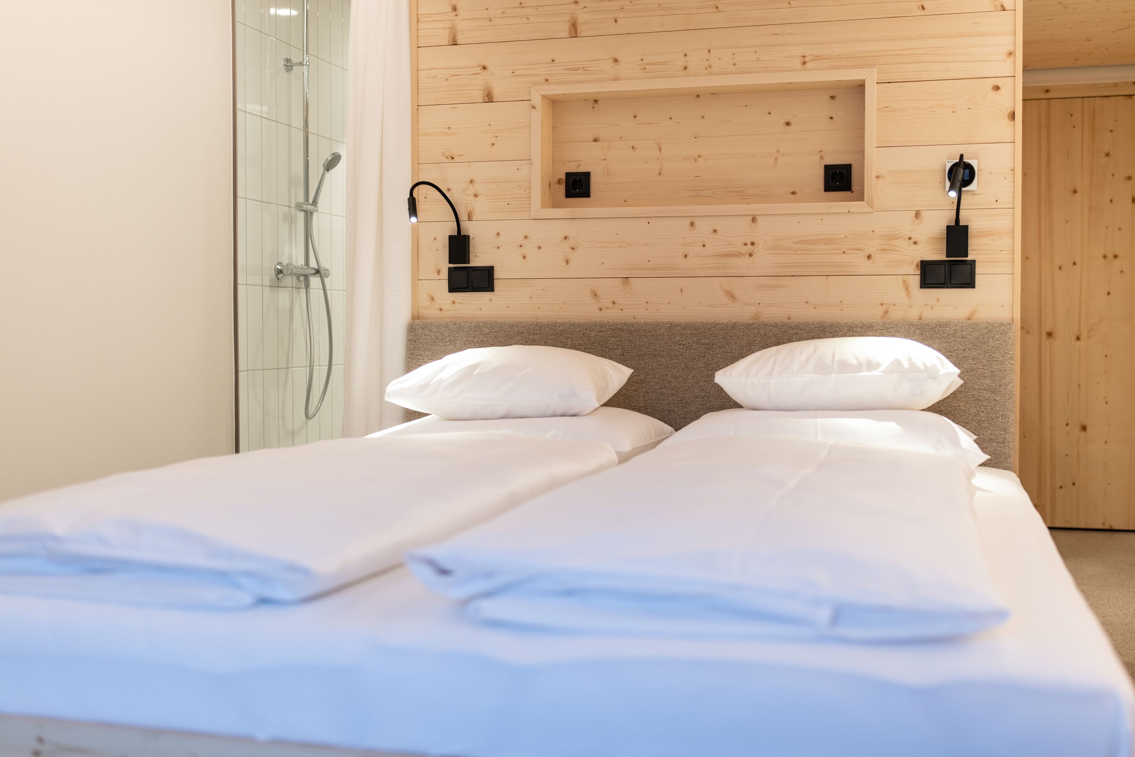 Quarto de hotel moderno com parede de madeira, box de vidro e cama com roupa de cama branca.