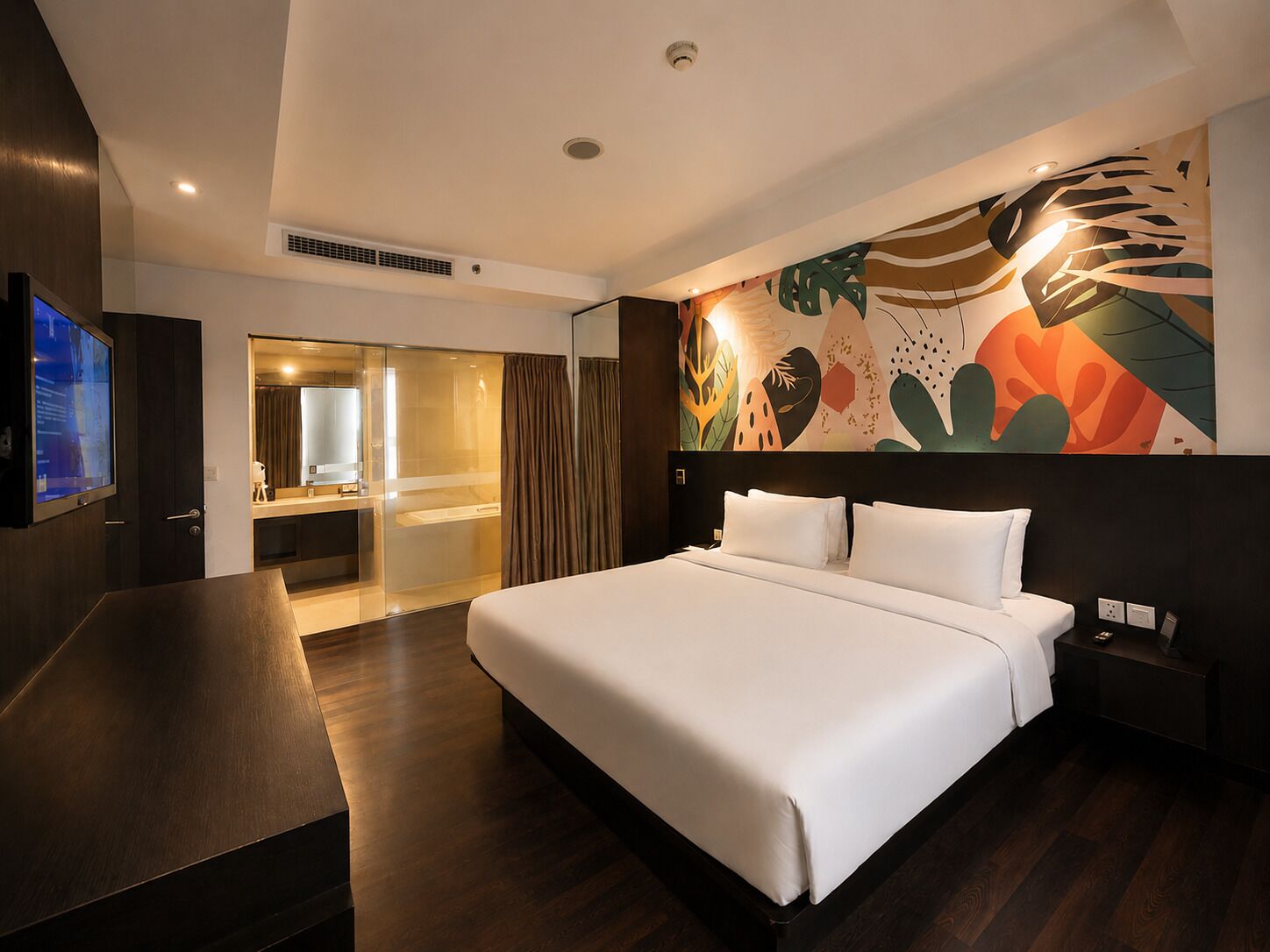 Quarto de hotel com uma cama grande, uma televisão, banheiro com porta de vidro e um mural abstrato e colorido na parede.
