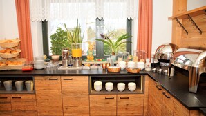 Buffet de café da manhã com diversos alimentos, utensílios, armários de madeira e uma janela com cortinas.