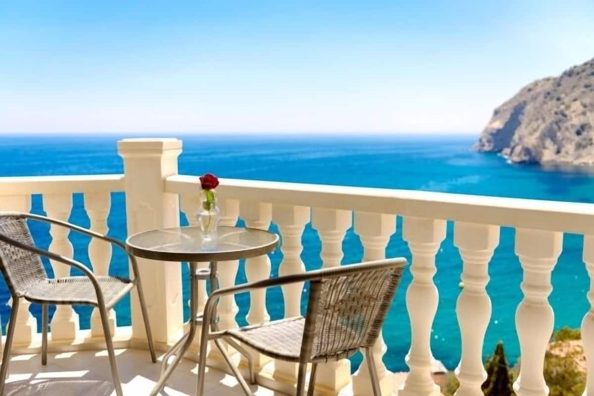 Varanda com duas cadeiras de vime e uma pequena mesa redonda, com vista para um mar azul límpido e um litoral rochoso.