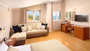 Quarto de hotel com sofá-cama, escrivaninha, cadeira, televisão e vista para o mar.