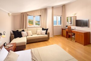 Quarto de hotel com sofá-cama, escrivaninha, cadeira, televisão e vista para o mar.