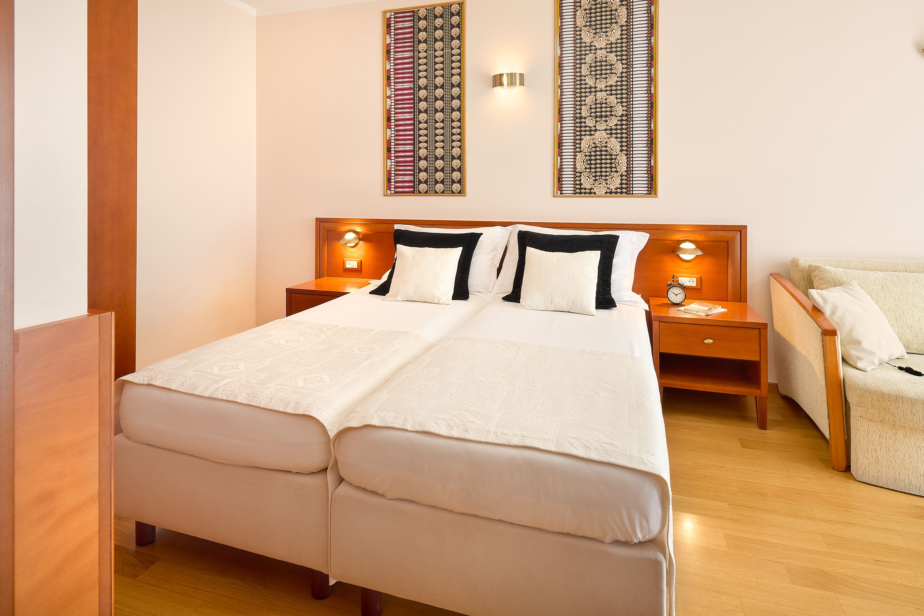 Uma cama de casal com cabeceira de madeira, duas mesas de cabeceira com abajures e um criado-mudo com relógio.