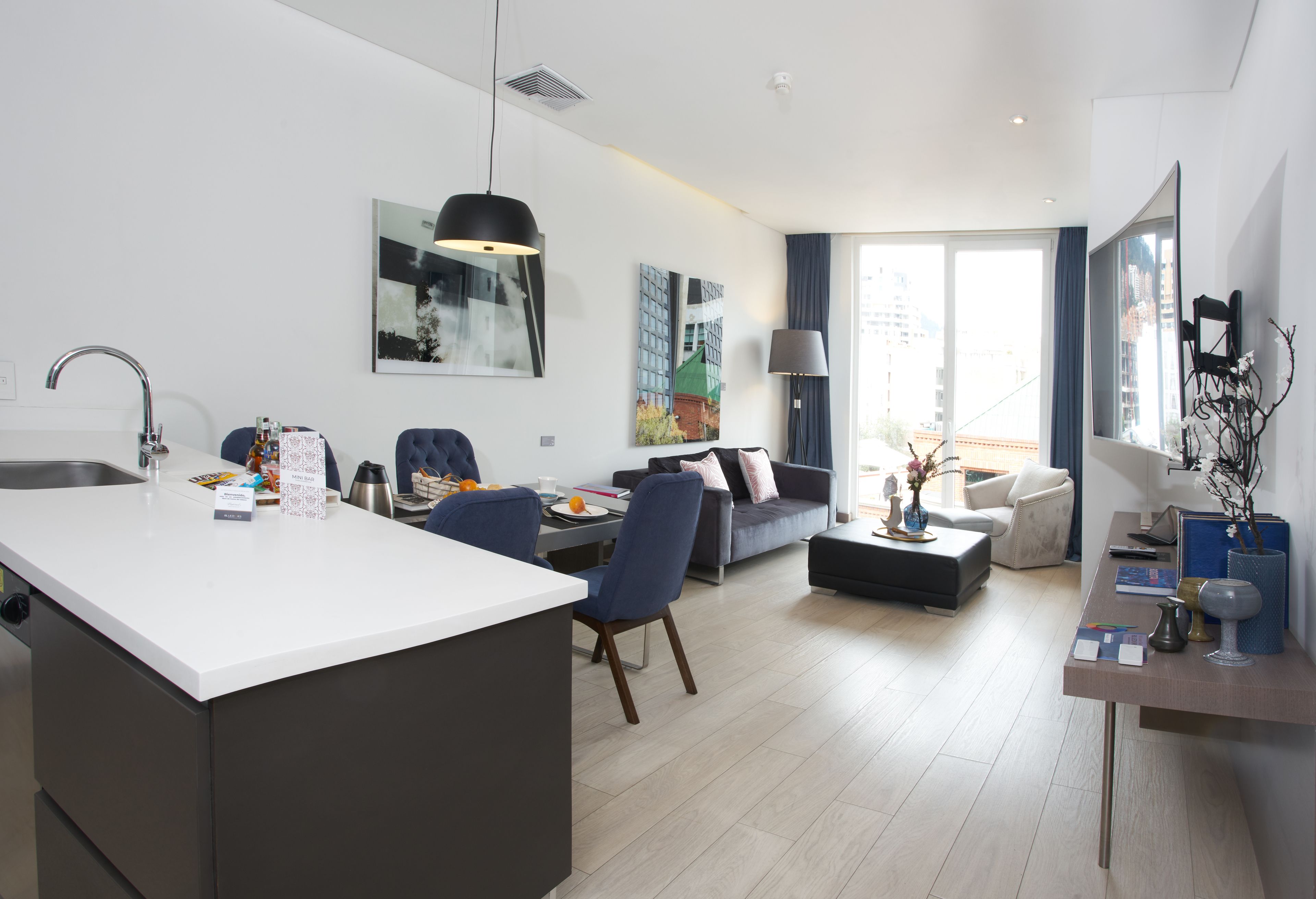 Quarto de hotel moderno com cozinha, espaço para refeições e área de estar com sofá e poltronas.