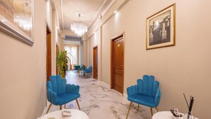 Corredor de hotel com piso de mármore, cadeiras estofadas em azul, um candelabro e obras de arte emolduradas.