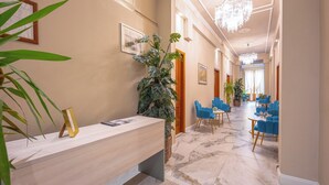 Um saguão de hotel com balcão de recepção, cadeiras azuis, lustres e obras de arte emolduradas nas paredes.