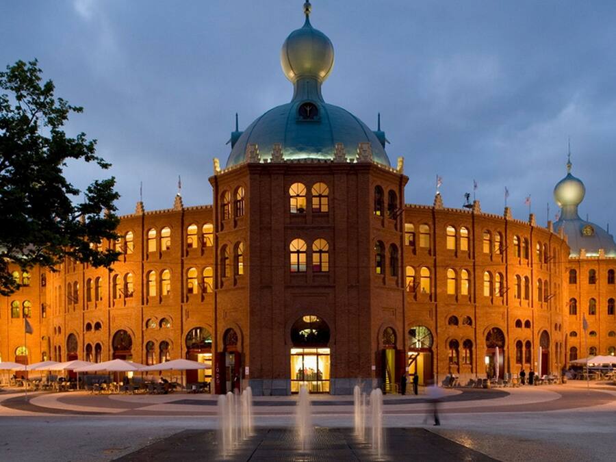 Um edifício grande e histórico, com uma cúpula central e várias cúpulas menores, rodeado por um pátio amplo com fontes e áreas de estar ao ar livre.