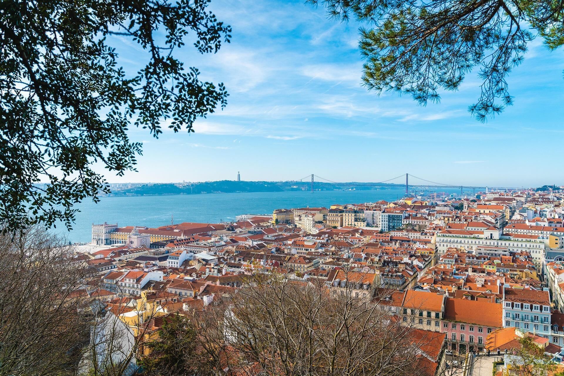 Vista panorâmica de uma cidade costeira, com uma ponte imponente ao longe, arquitetura urbana densa e céu claro.