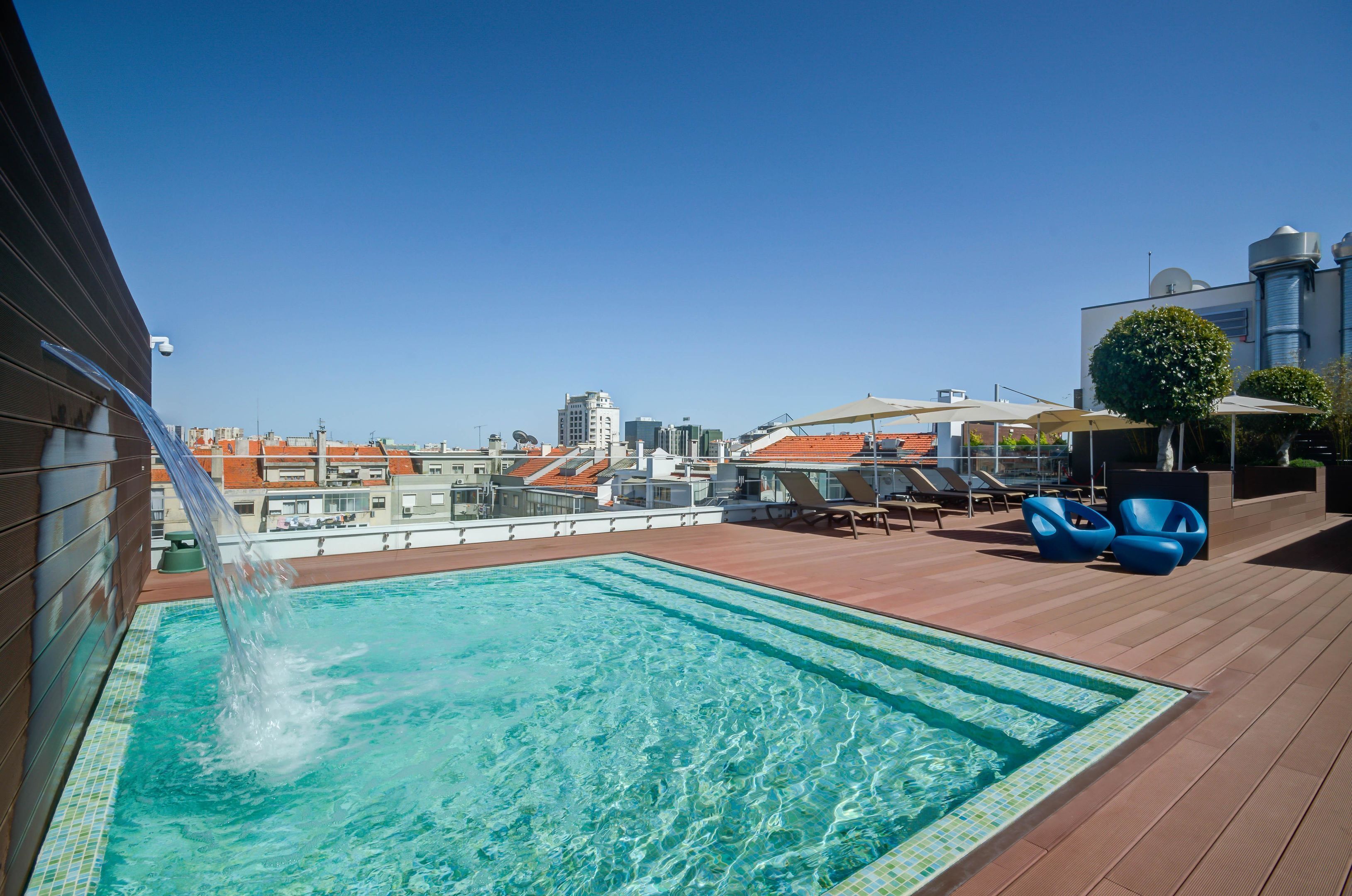 Piscina no terraço com um recurso aquático, cadeiras de descanso e vista para a paisagem urbana.