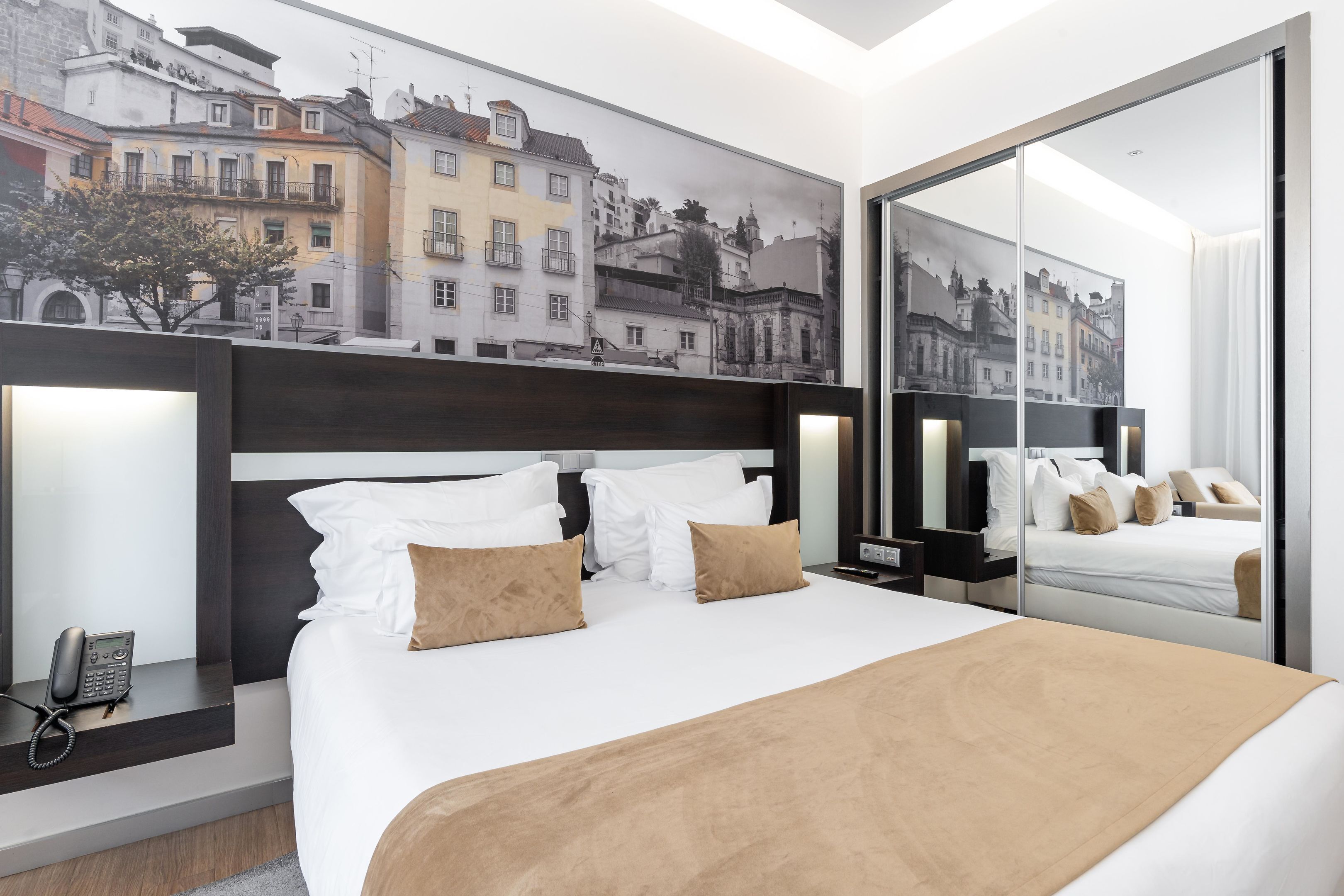 Quarto de hotel moderno com uma cama grande, telefone ao lado e um mural com vista para a cidade na parede.