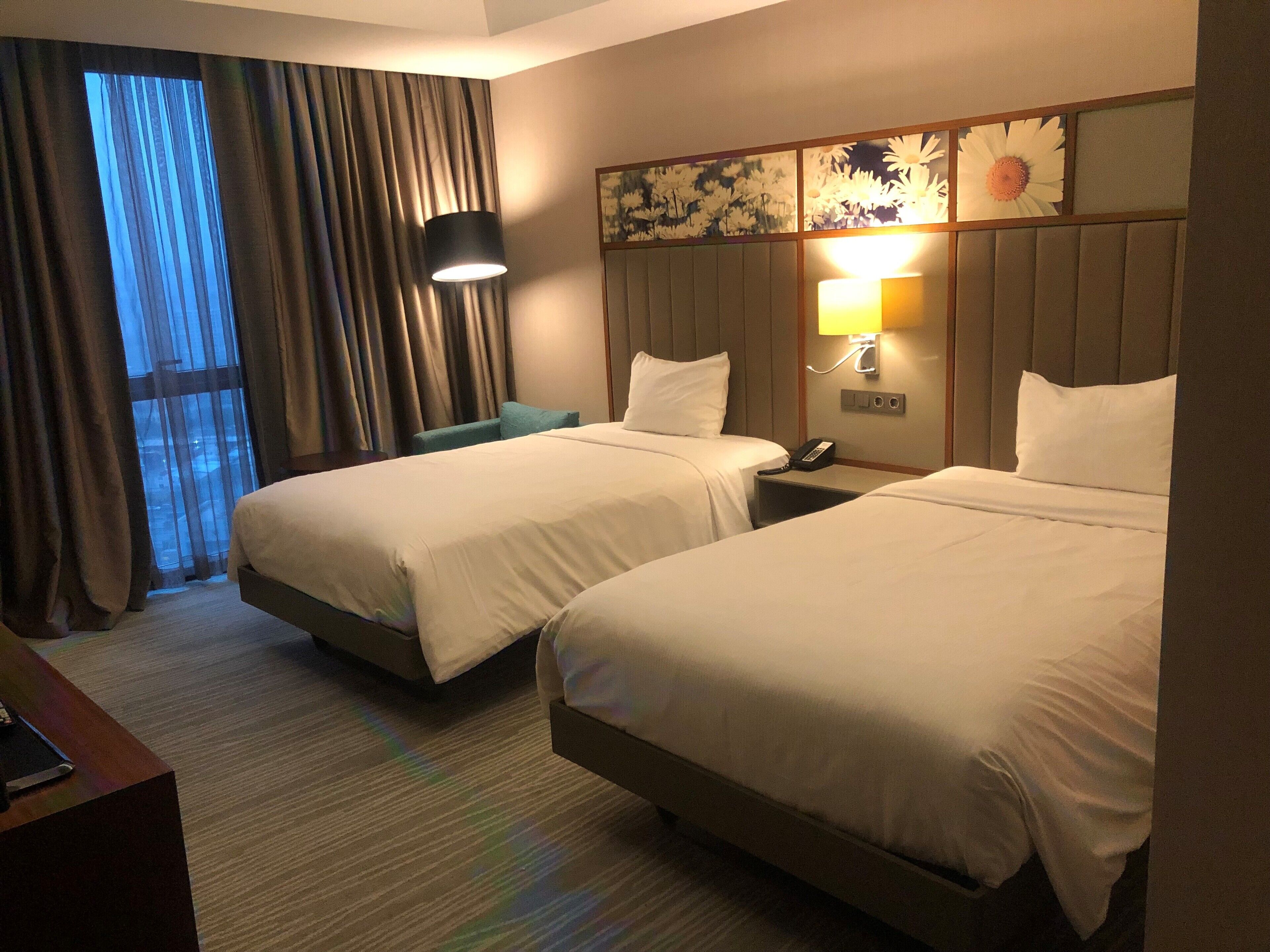 Quarto de hotel com duas camas, uma escrivaninha, uma cadeira, um abajur e uma janela com cortinas.