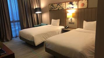 Quarto de hotel com duas camas, uma escrivaninha, uma cadeira, um abajur e uma janela com cortinas.