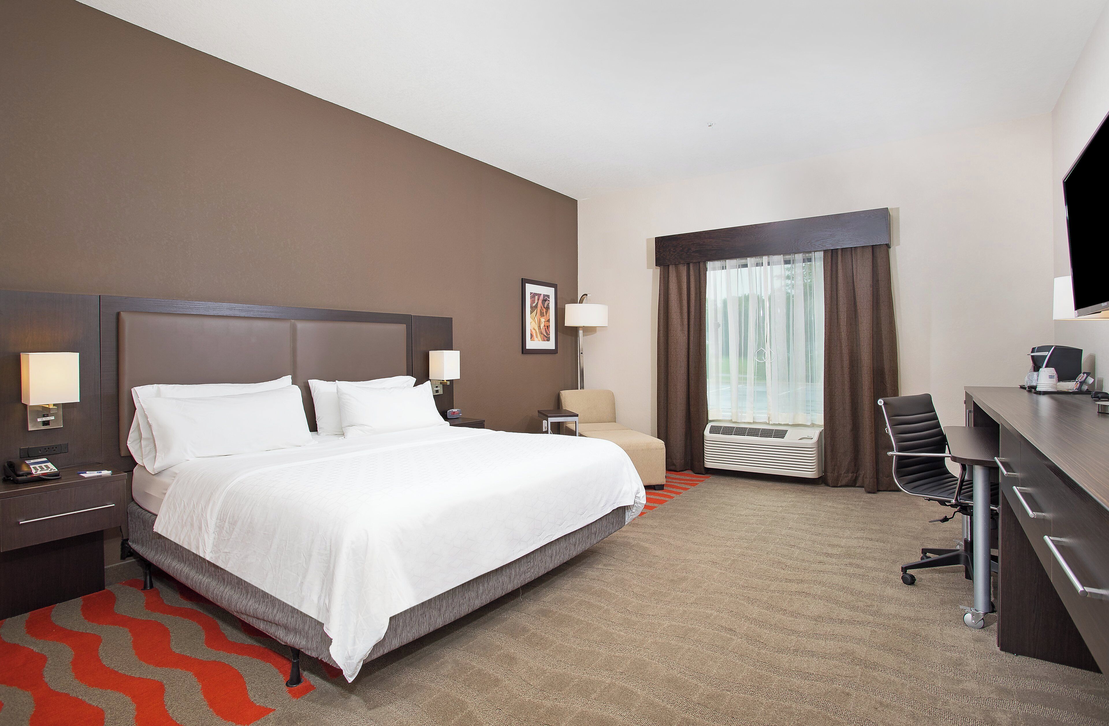 Quarto de hotel com cama grande, mesas de cabeceira, escrivaninha com cadeira, janela com cortinas e TV fixada na parede.