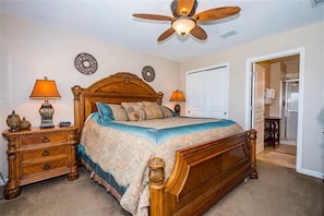 Quarto com cama de madeira, duas mesas de cabeceira com abajures, um ventilador de teto e uma porta que dá para o banheiro.