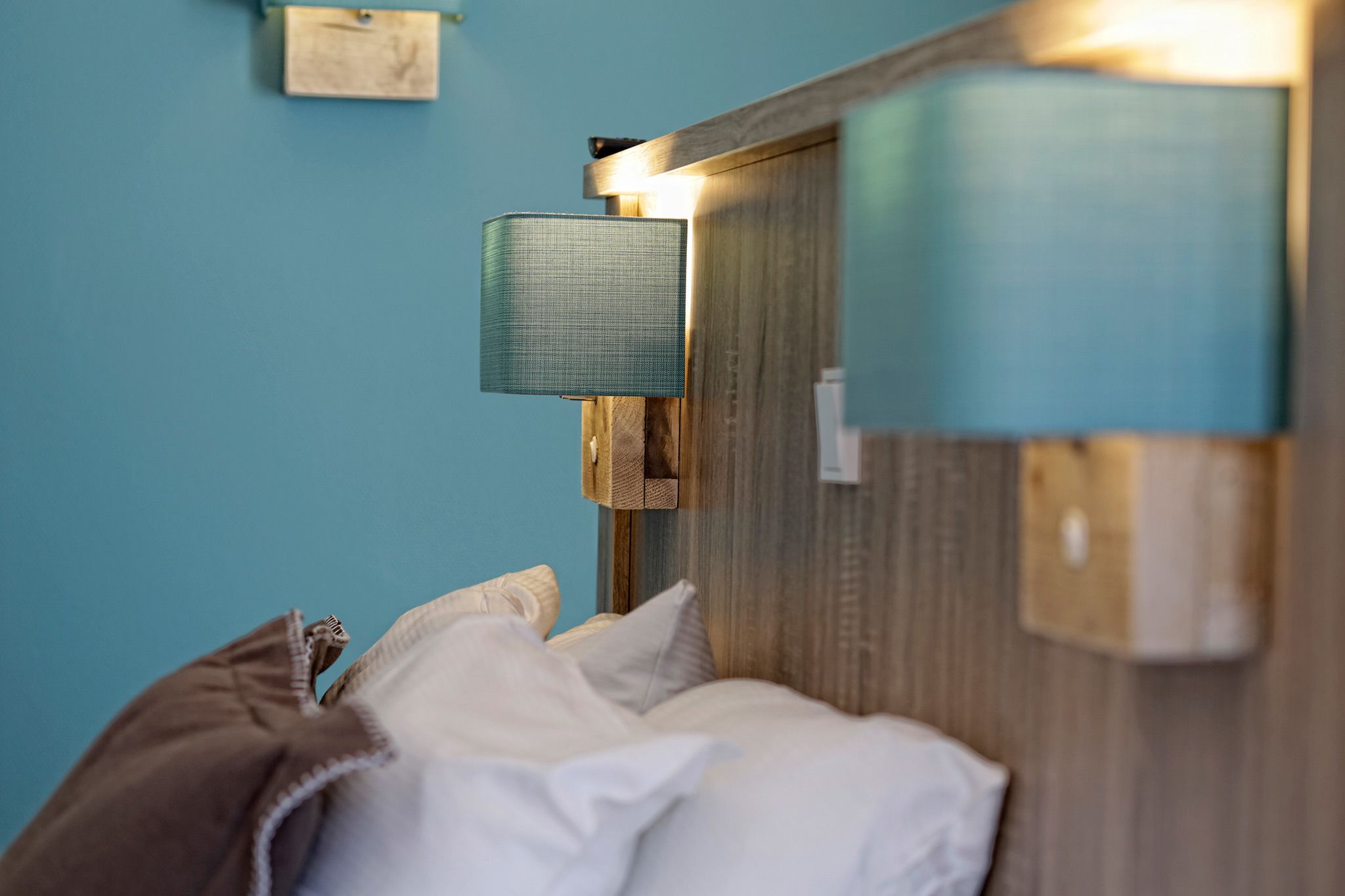 Uma cama bem arrumada com roupa de cama branca e castanha, uma cabeceira de madeira e uma luminária fixada na parede.