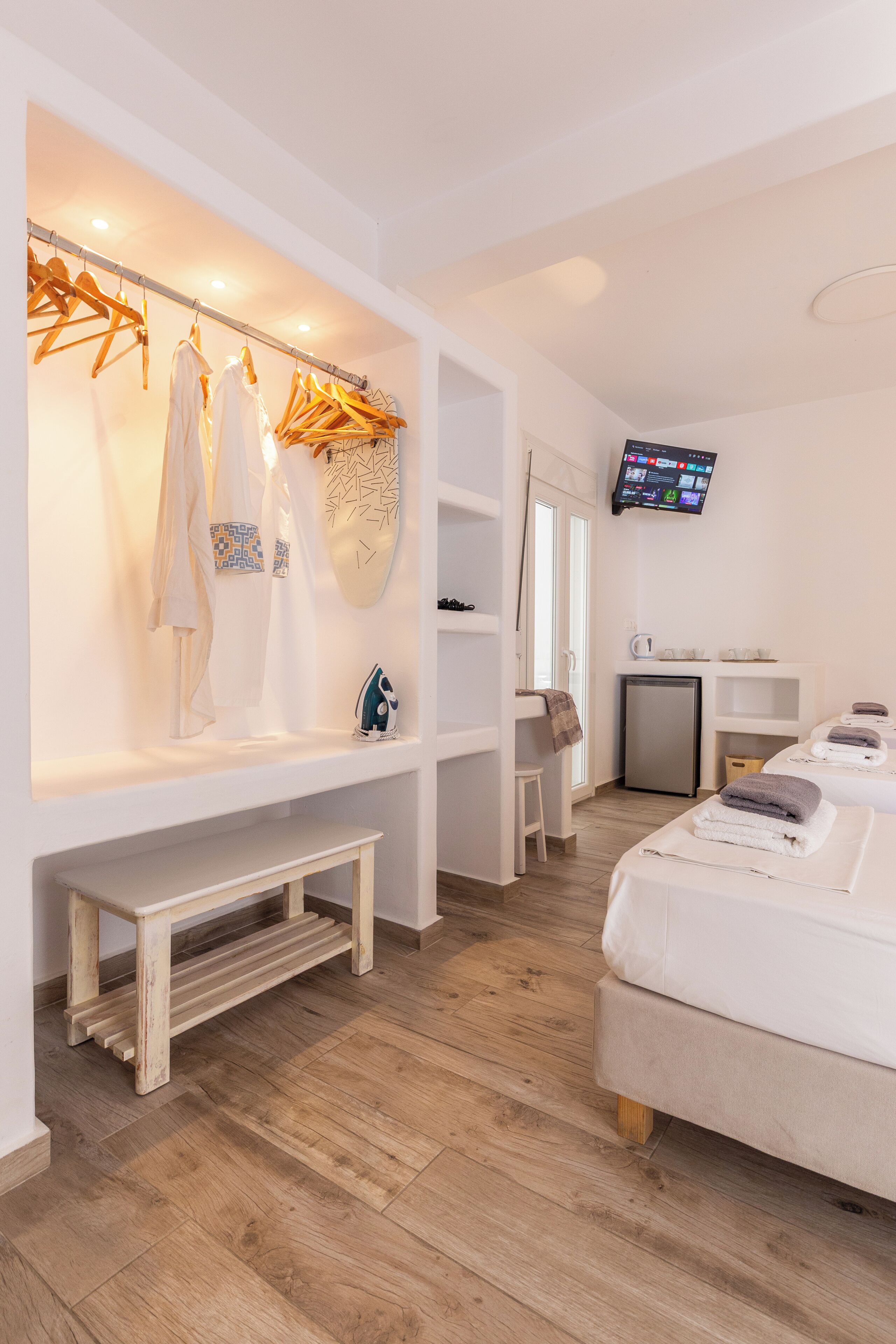 Quarto de hotel com cama, televisão na parede, um banco de madeira e um cabideiro com cabides.