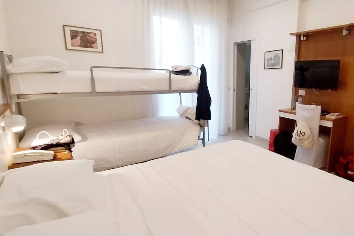 Quarto de hotel com beliches, uma mesa com cadeira, uma televisão fixada em um painel de madeira e um guarda-roupa.