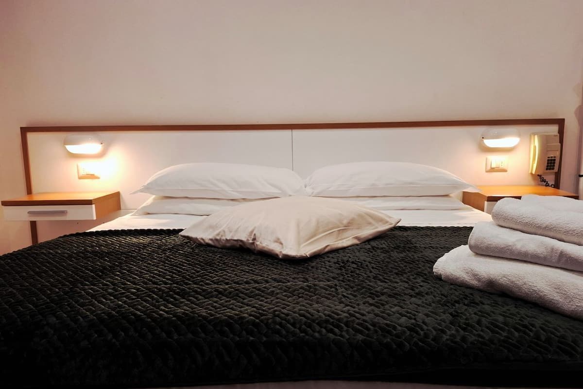 Cama bem arrumada com um cobertor escuro texturizado, travesseiros brancos e toalhas dobradas ao lado direito. Existem duas mesas de cabeceira com luminárias de cada lado da cama.