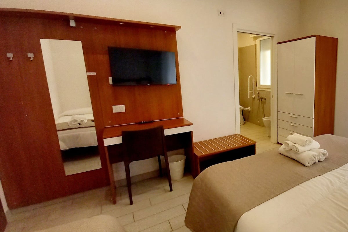 Quarto de hotel com cama, escrivaninha, cadeira, televisão e guarda-roupa.