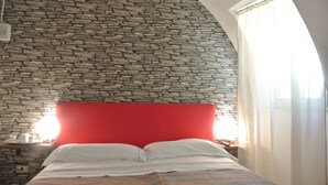 Quarto com parede de pedra, cabeceira vermelha, cama com roupa de cama branca e janela com cortinas.
