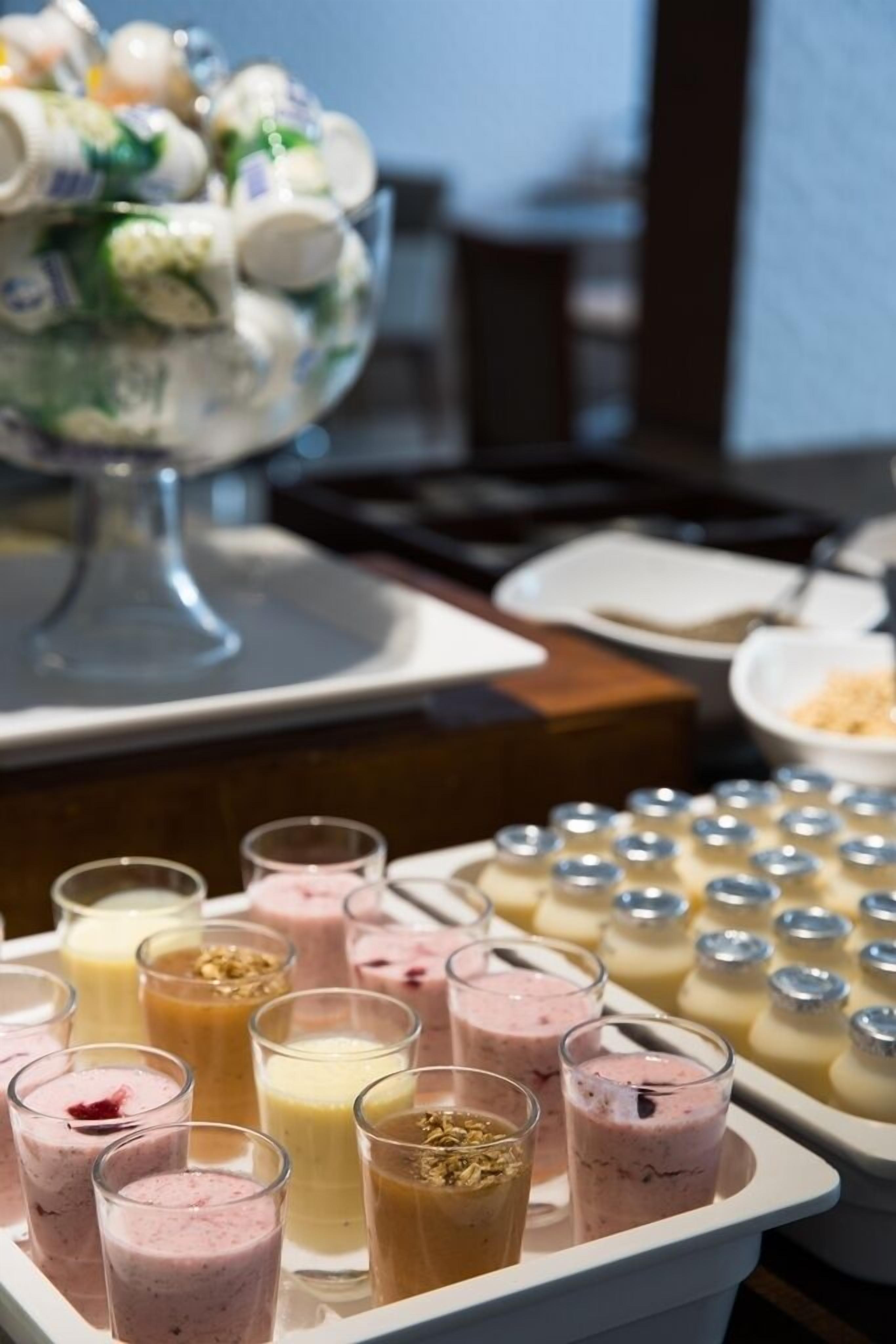 Mesa de buffet com diversos alimentos, incluindo uma saladeira, uma bandeja com pequenos copos de bebidas coloridas e um recipiente com potinhos de iogurte.
