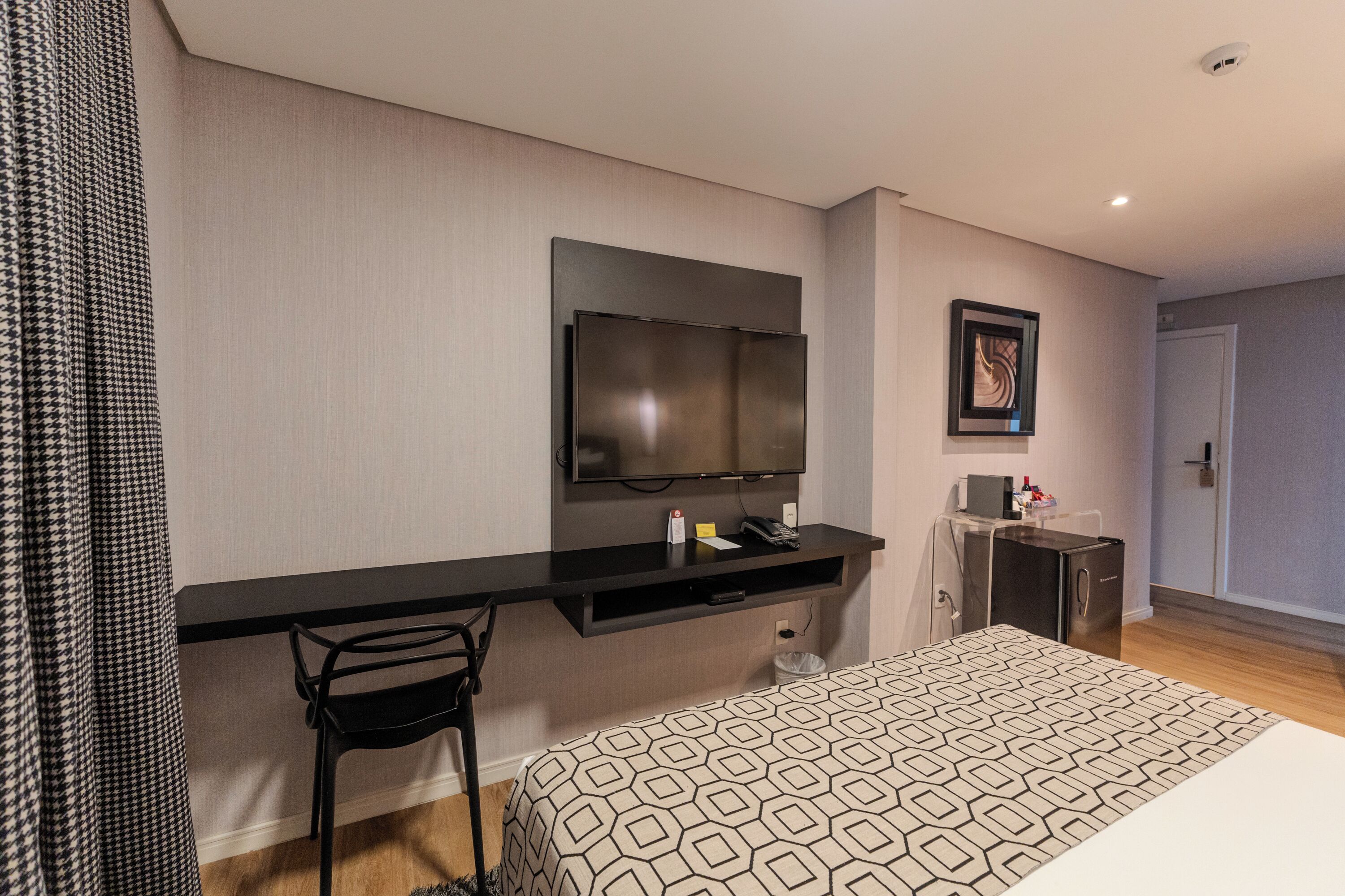 Quarto de hotel com uma cama, uma escrivaninha com cadeira, TV fixada na parede, mini-frigorífico e um cesto de lixo.