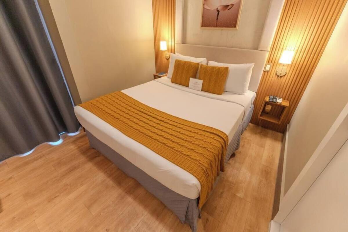 Cama bem arrumada com manta listrada em branco e castanho, duas almofadas e cabeceira de madeira. Há duas mesas de cabeceira com abajures, e um luminário fixado na parede acima da cama.