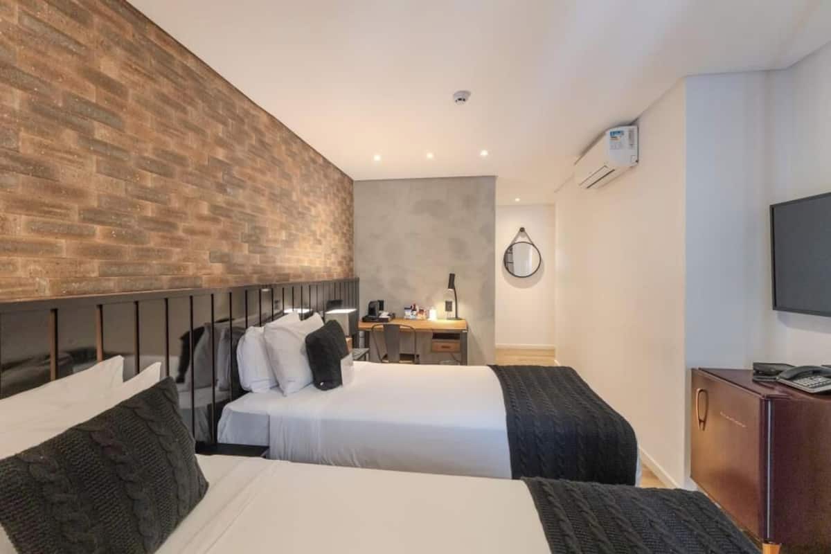 Quarto de hotel com duas camas, uma escrivaninha, uma cadeira, uma televisão e uma parede de tijolos.