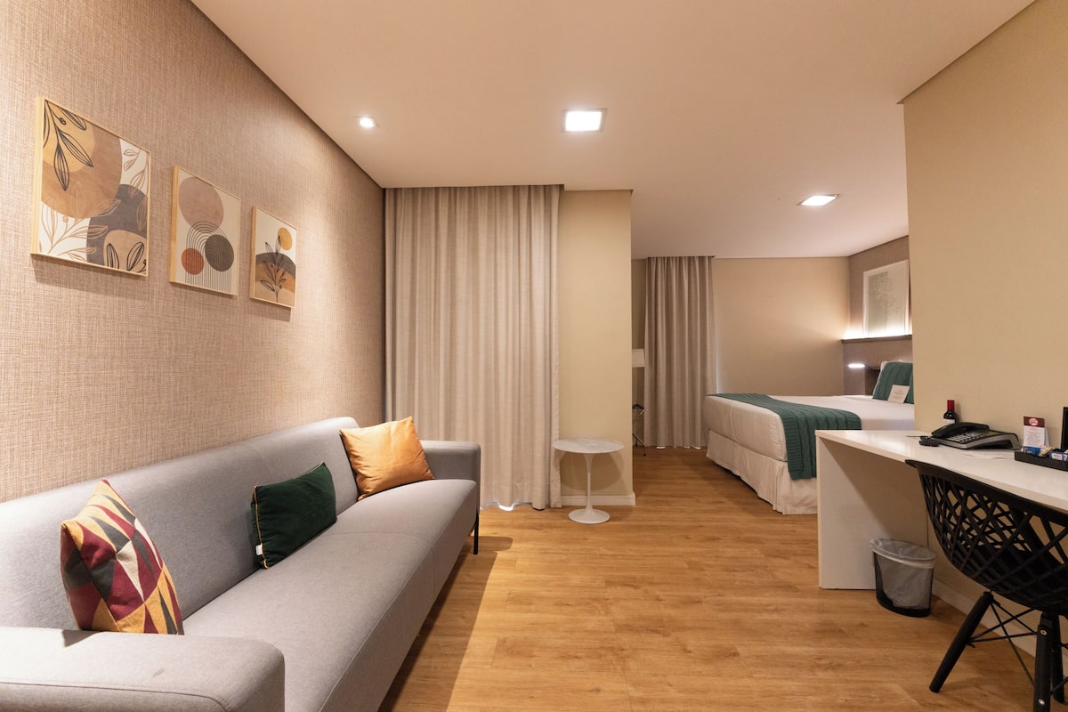 Quarto de hotel moderno com um sofá cinza, uma mesa redonda pequena, uma cama com manta verde e uma escrivaninha com cadeira.