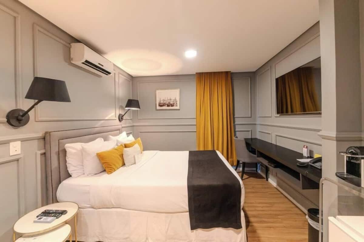 Quarto de hotel com cama, mesas de cabeceira, escrivaninha e janela com cortinas.