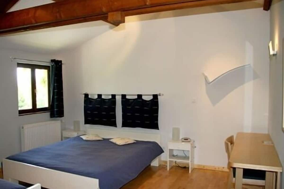 Quarto com cama, mesinha, cadeira, janela com cortinas e radiador.