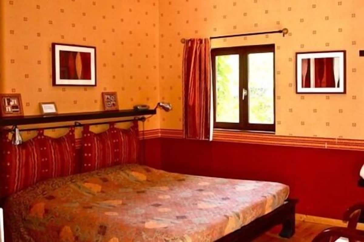 Quarto com cama, duas janelas com cortinas, um prateleira com objetos e parede estampada.