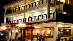 Hotel com vários andares, varanda no andar superior, restaurante no nível da rua com mesas ao ar livre e fachada bem iluminada.