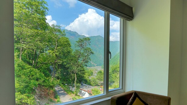 Quarto com sofá, mesa de vidro, janela com vista para vegetação e montanhas.