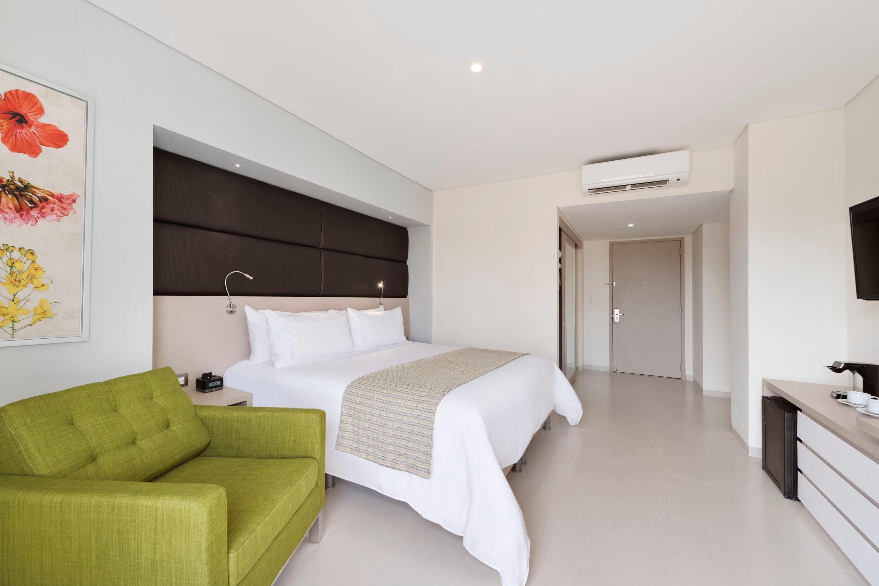 Quarto de hotel moderno com uma cama grande, um sofá verde, uma televisão de tela plana e um quadro na parede.
