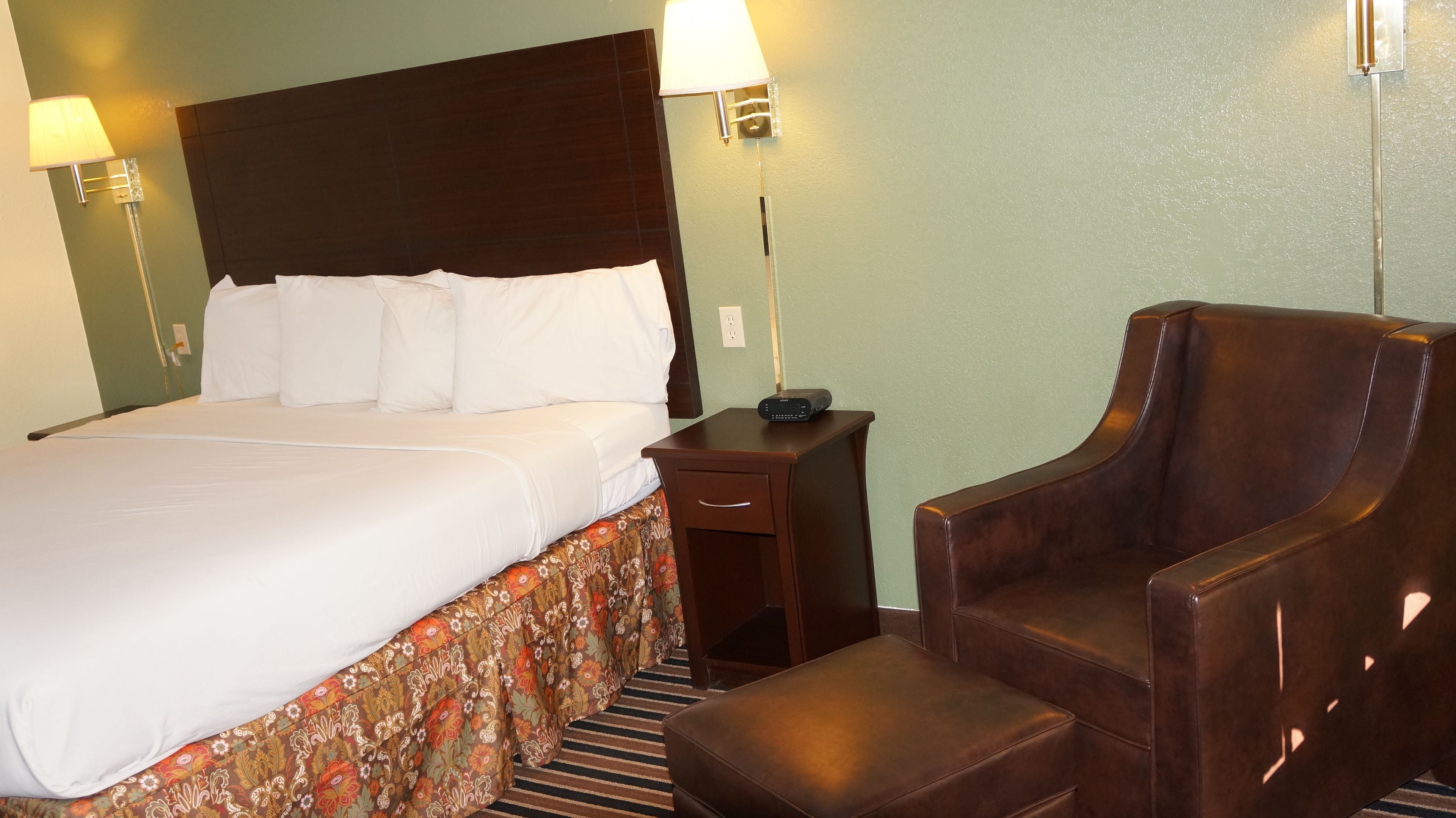 Quarto de hotel com cama, uma cadeira, mesa de cabeceira e duas luminárias de parede.