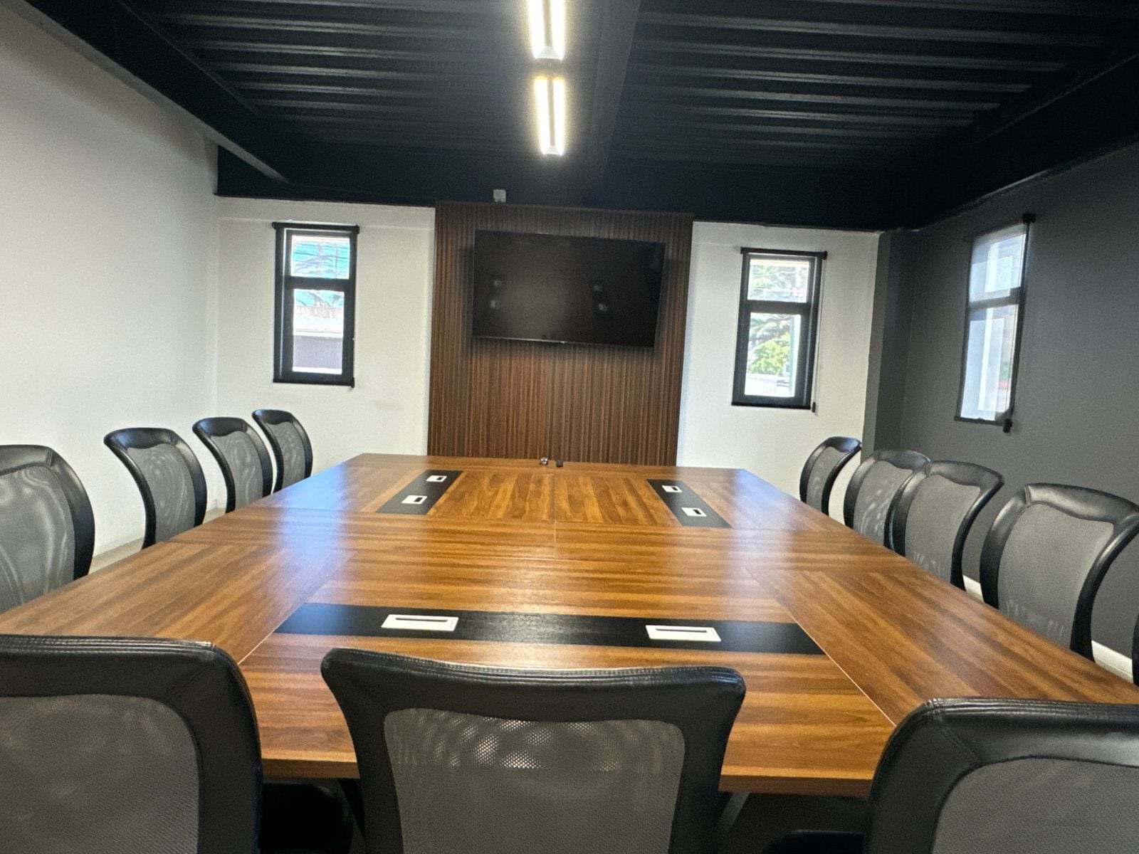 Uma sala de conferências com uma longa mesa de madeira, cadeiras pretas, uma TV de tela plana e janelas com persianas.