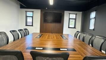 Uma sala de conferências com uma longa mesa de madeira, cadeiras pretas, uma TV de tela plana e janelas com persianas.
