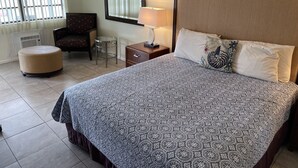 Quarto de hotel com uma cama grande, uma cadeira, uma mesinha e uma luminária.