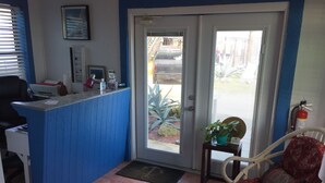 Uma área de recepção com balcão azul, uma cadeira, uma pequena mesa com uma planta e uma porta que dá para o exterior.