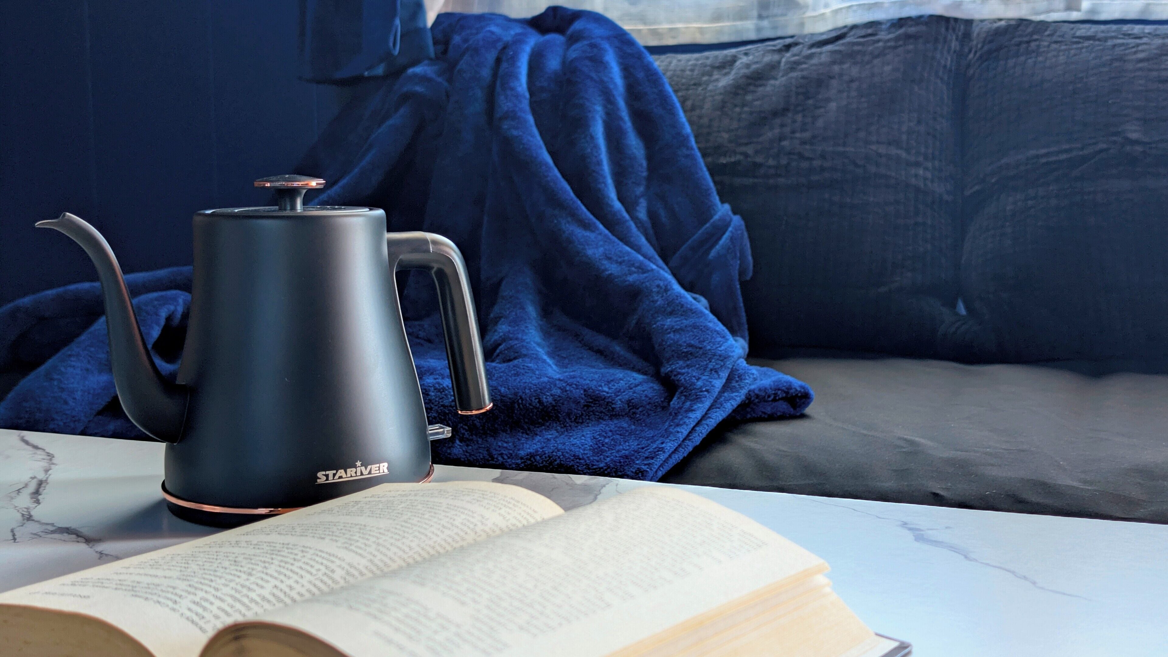 Uma chaleira elétrica preta sobre uma mesa, com um livro aberto, um cobertor azul e um sof á cinzento ao fundo.