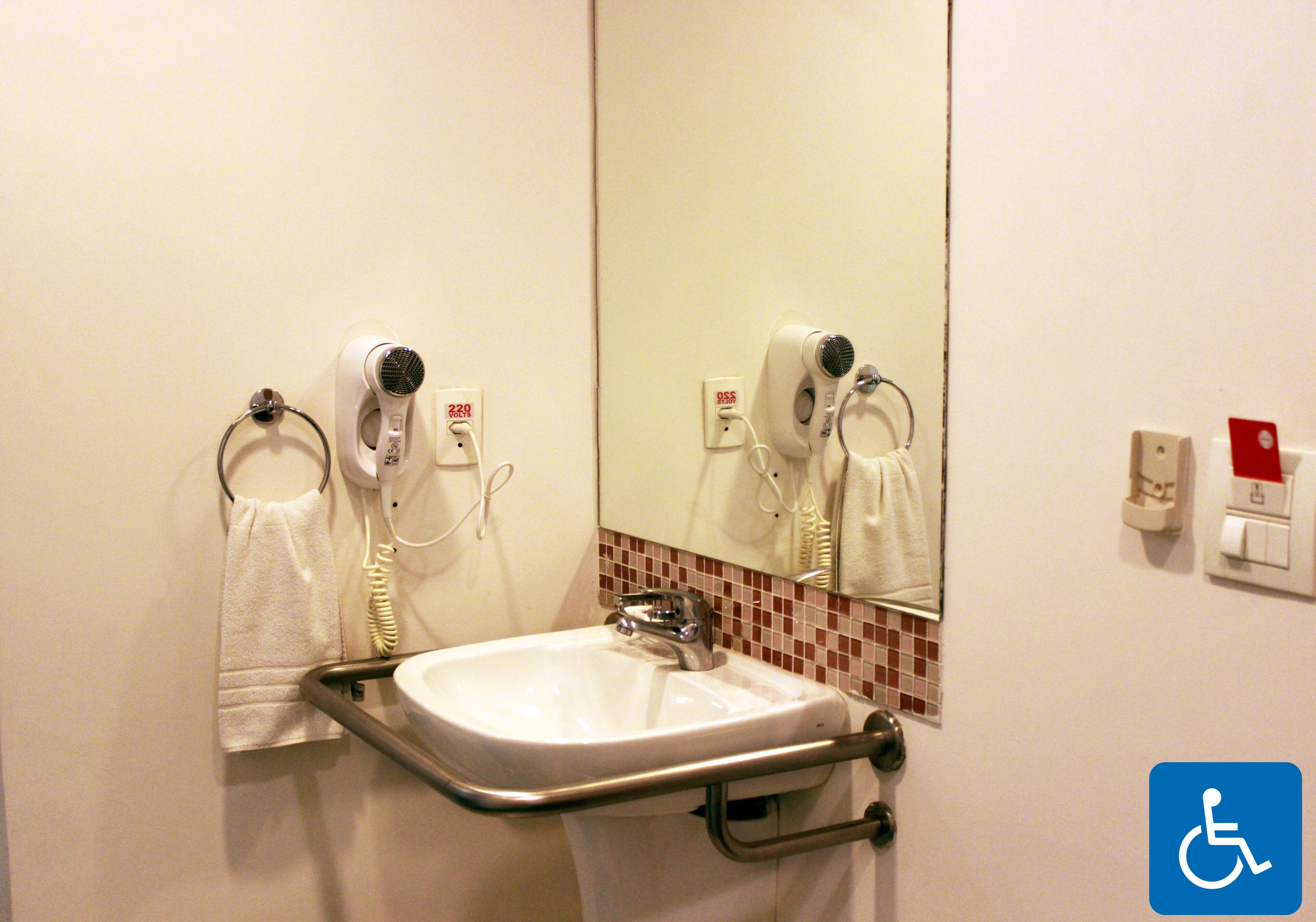 Casa de banho com lavatório, dois secadores de cabelo, espelho e barra de apoio.